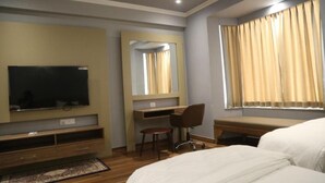 Quarto de hotel com cama, escrivaninha, cadeira, televisão e uma janela grande com cortinas.