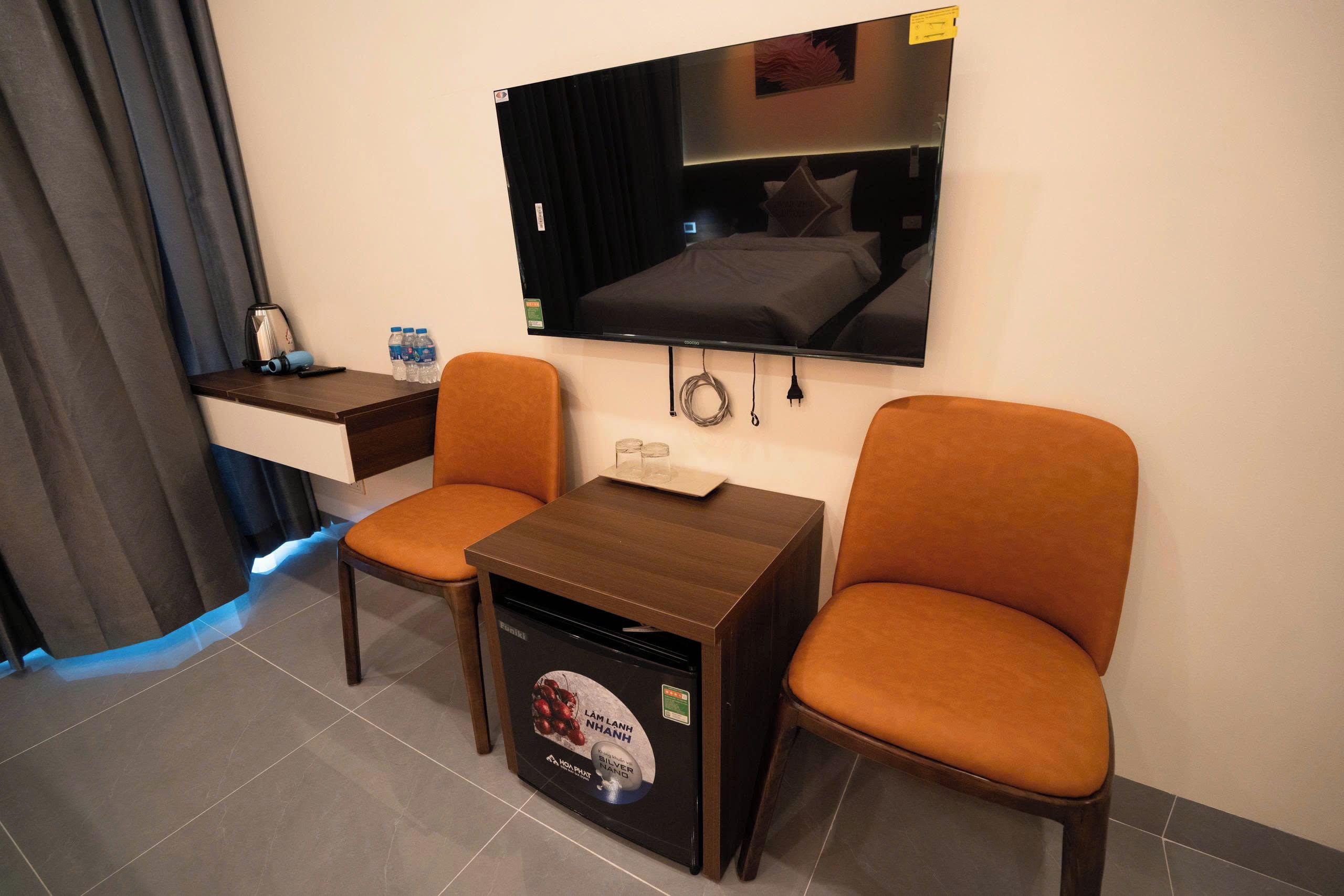 Quarto de hotel com cama, televisão na parede, pequena mesa com cadeira e minibar.