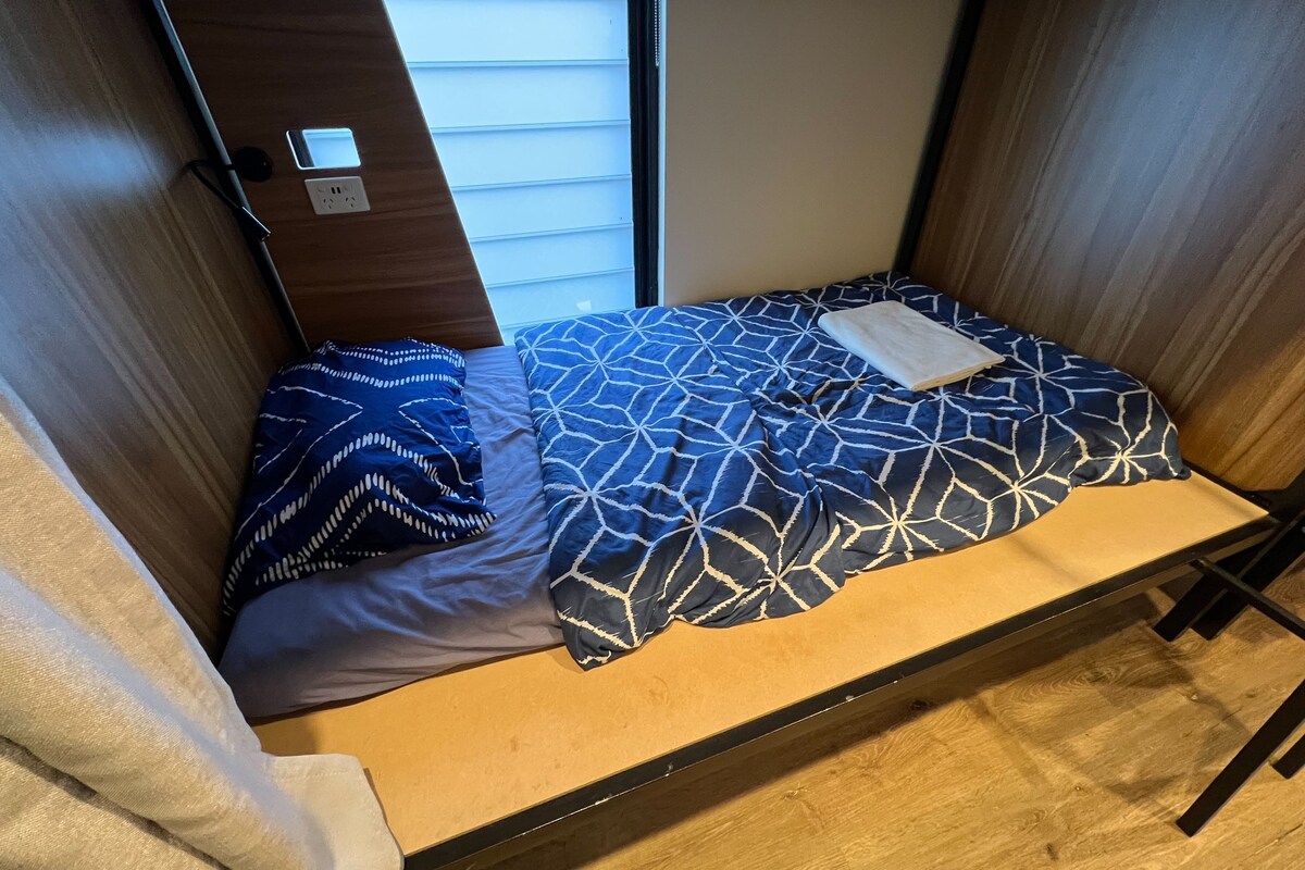 Uma cama de solteiro com colcha e almofadas estampadas, cabeceira de madeira e uma tomada.