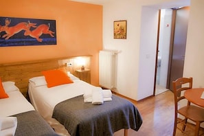 Quarto de hotel com duas camas, cabeceira de madeira, radiador, quadro e porta que dá para outro cômodo.