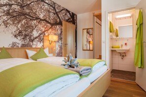 Quarto de hotel com duas camas, uma janela grande com vista para uma árvore, banheiro com pia e espelho, e porta que dá para outro quarto.