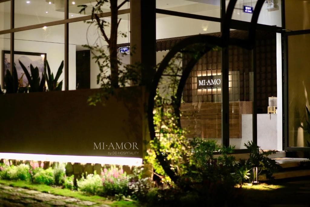 Exterior moderno de um hotel com uma grande entrada em arco, uma placa bem iluminada com a inscrição 'MI-AMOR by DD Hospitality' e um jardim com plantas e um caminho.