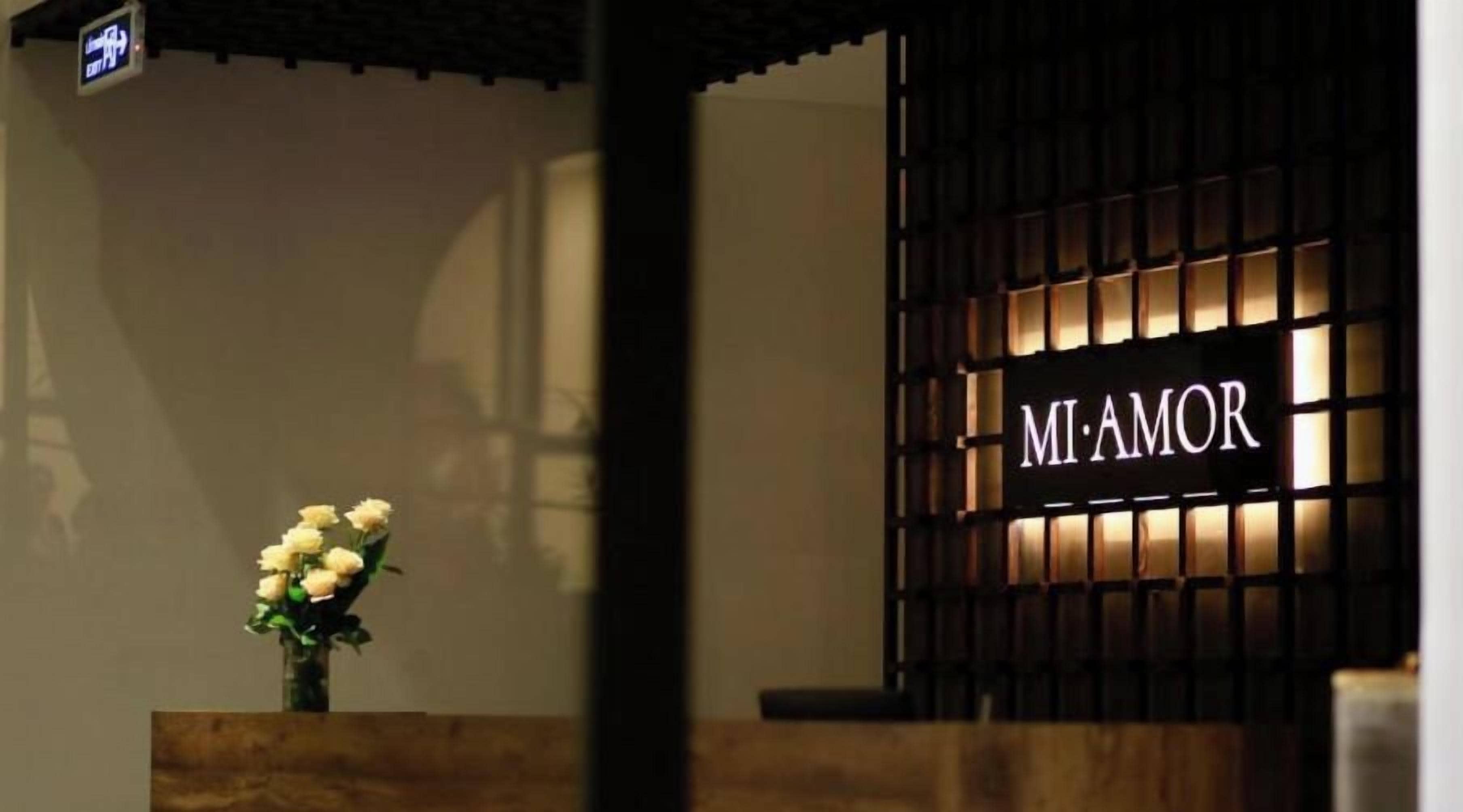 Balcão de recepção com uma placa que diz 'MI-AMOR' e um arranjo floral à esquerda.