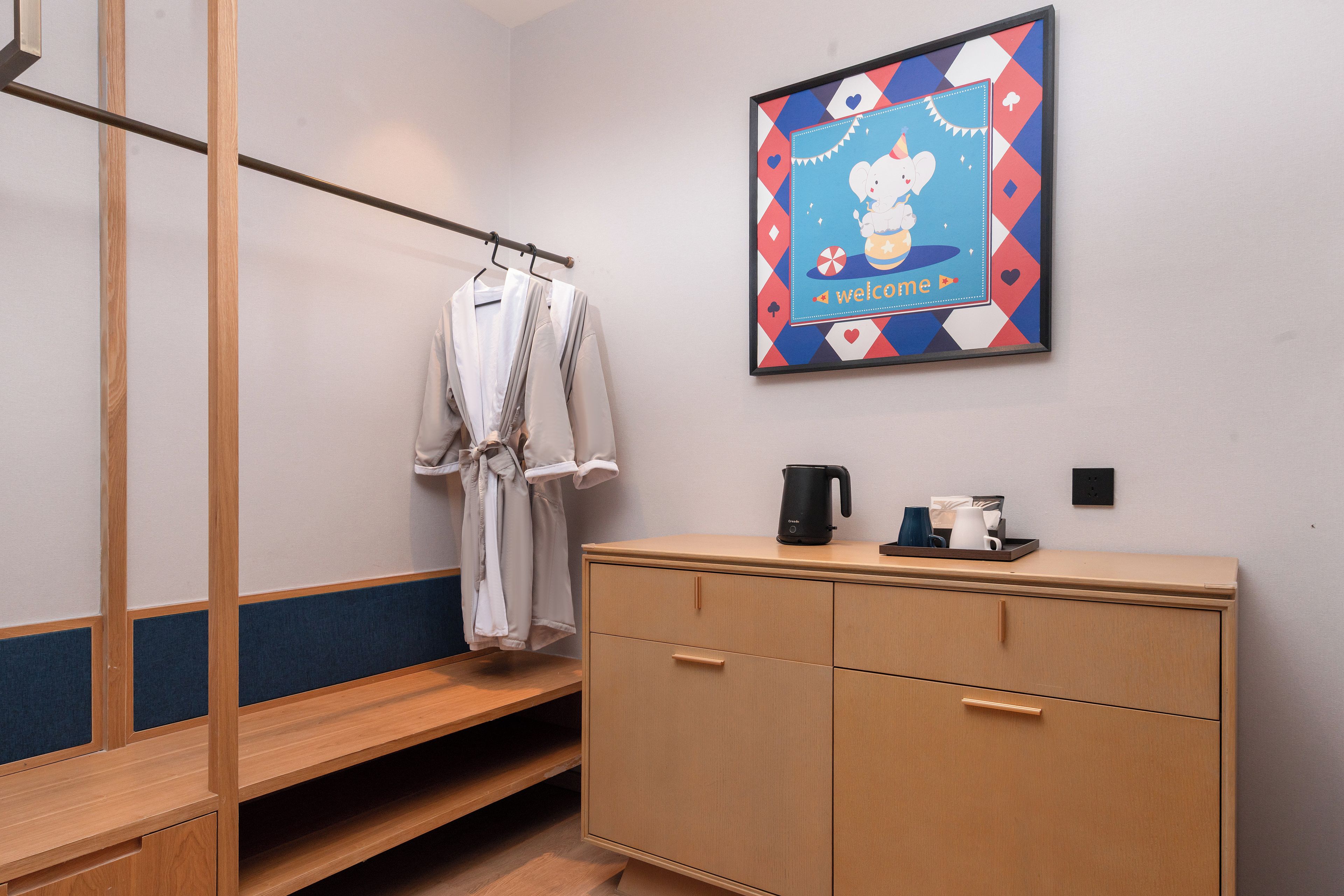 Quarto de hotel com um banco de madeira, guarda-roupa, cabides com roupões e um quadro na parede.
