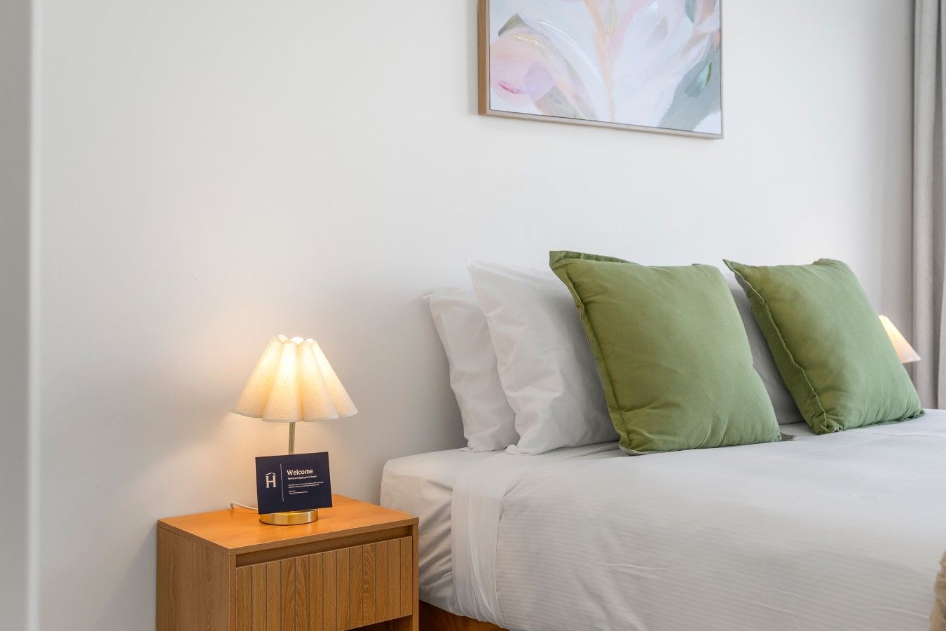 Uma cama bem arrumada com almofadas brancas e verdes, um abajur e uma placa de boas-vindas em uma mesa de cabeceira de madeira.