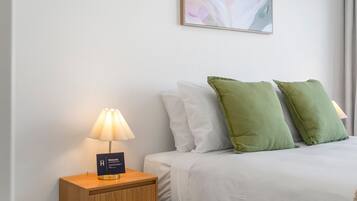 Uma cama bem arrumada com almofadas brancas e verdes, um abajur e uma placa de boas-vindas em uma mesa de cabeceira de madeira.