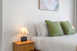 Uma cama bem arrumada com almofadas brancas e verdes, um abajur e uma placa de boas-vindas em uma mesa de cabeceira de madeira.