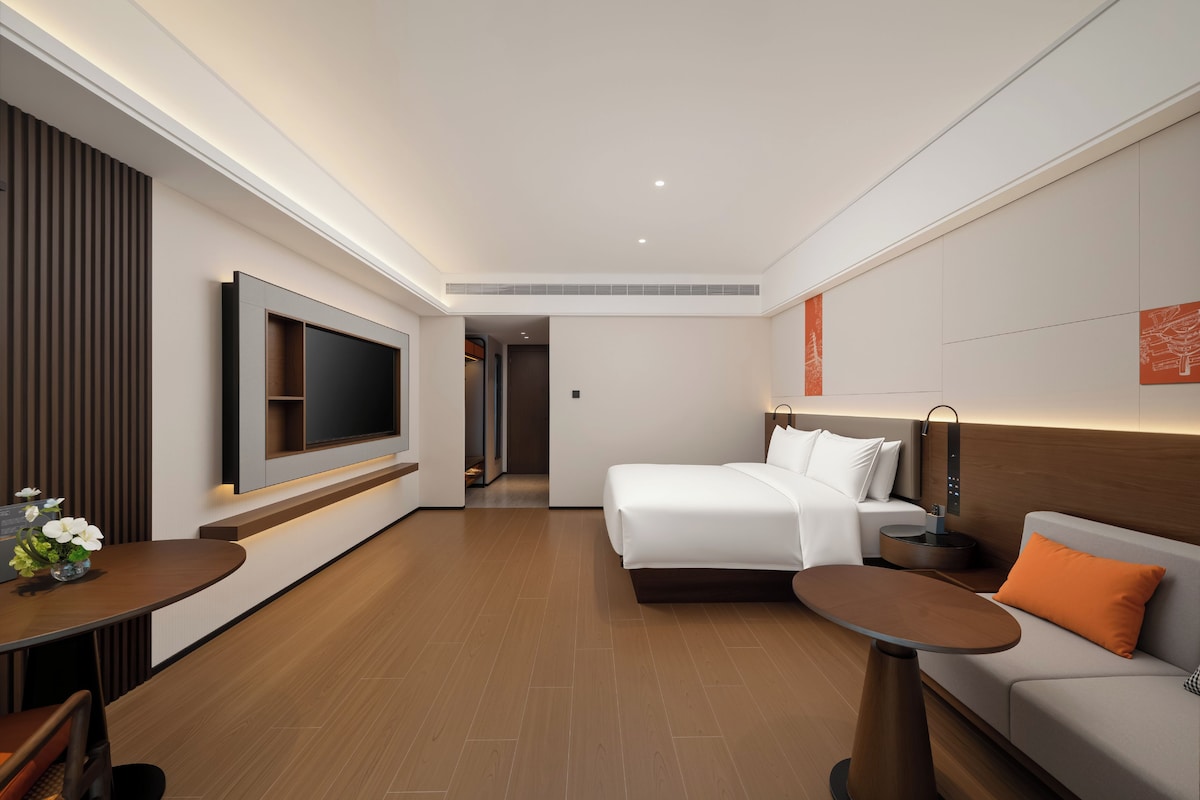 Quarto de hotel moderno com televisão de tela plana, cama, sofá, mesinha e um vaso decorativo.