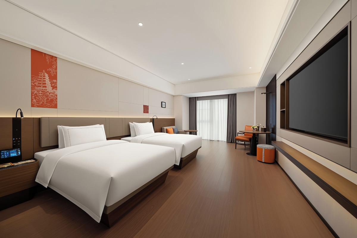 Quarto de hotel com duas camas, uma escrivaninha, uma cadeira, televisão e uma janela grande.