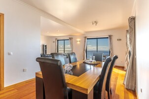 Uma sala de jantar com uma mesa de madeira, cadeiras de couro e vista para o mar através de grandes janelas.