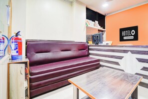 Área de recepção de hotel com balcão, um sofá roxo, uma mesa pequena e um extintor de incêndio.