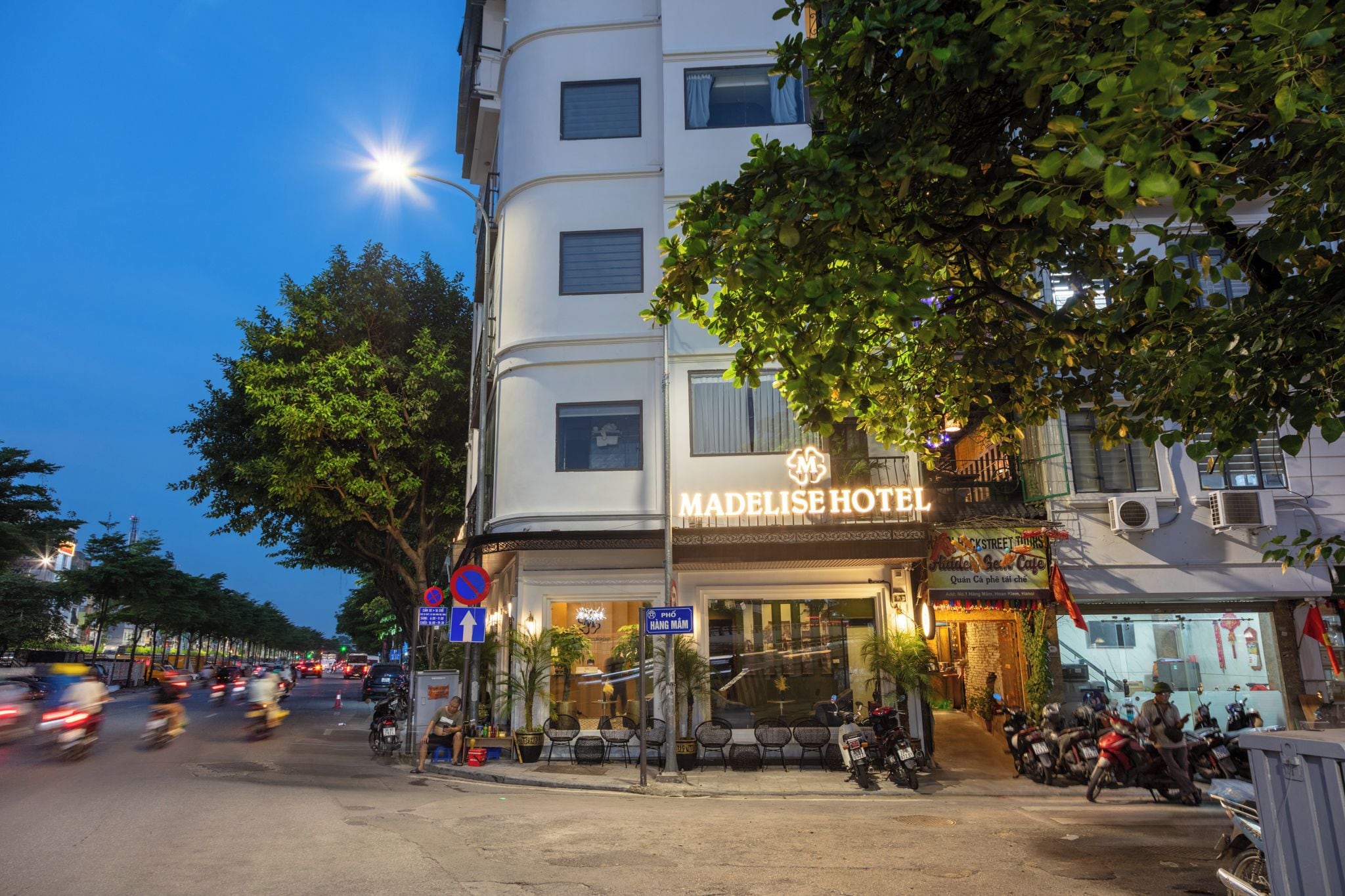 Hotel com placa indicando 'MADELIS HOTEL' e rua em frente com motocicletas estacionadas e trânsito.