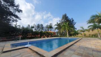 Piscina com deck ladrilhado, cercada por palmeiras e um edifício com telhado vermelho.