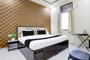 Quarto de hotel com cama, mesa de cabeceira, planta em vaso, escrivaninha com cadeira e janela com cortinas.