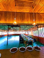 Um espaço de refeições interno com mesas e cadeiras, uma mesa longa com pratos e tigelas, e luminárias pendentes.