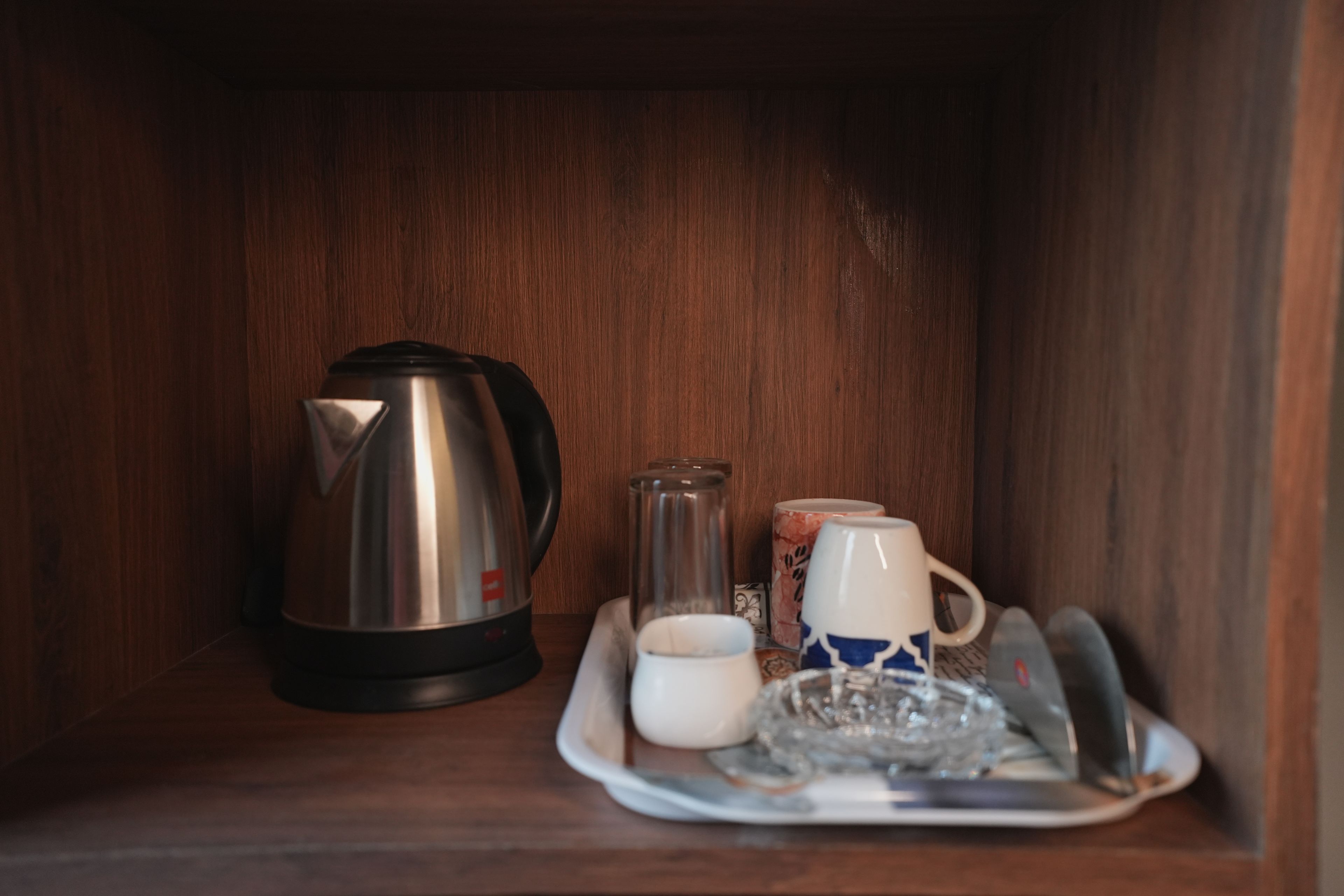 Um armário de madeira com uma chaleira de aço inoxidável, um jarro de vidro, uma caneca branca e uma bandeja com um copo e uma colher.
