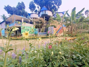 Um jardim com diversas flores e plantas, um edifício com telhado azul e uma escadaria que leva a uma varanda.