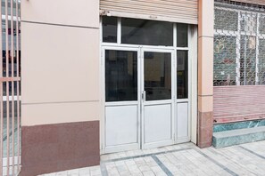 Entrada de edifício com portas duplas, portão de metal e pavimento ladrilhado.