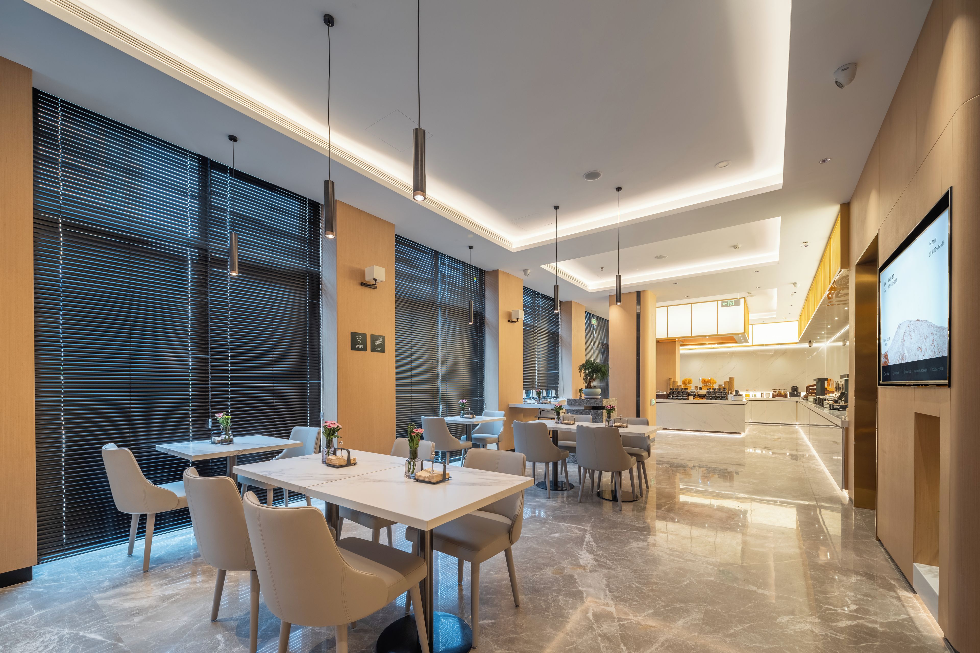 Um saguão de hotel moderno com área de refeições, cozinha ao fundo e uma tela grande exibindo informações.