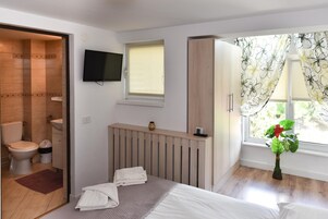 Quarto com cama, televisão, janela com cortinas, radiador e banheiro com vaso sanitário e pia.