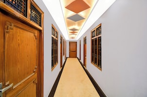 Um corredor com portas e janelas de madeira, piso ladrilhado e teto com luminárias geométricas.