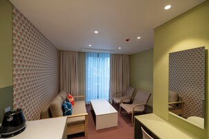 Quarto de hotel com cama, escrivaninha, duas cadeiras, máquina de café e uma janela grande com cortinas.