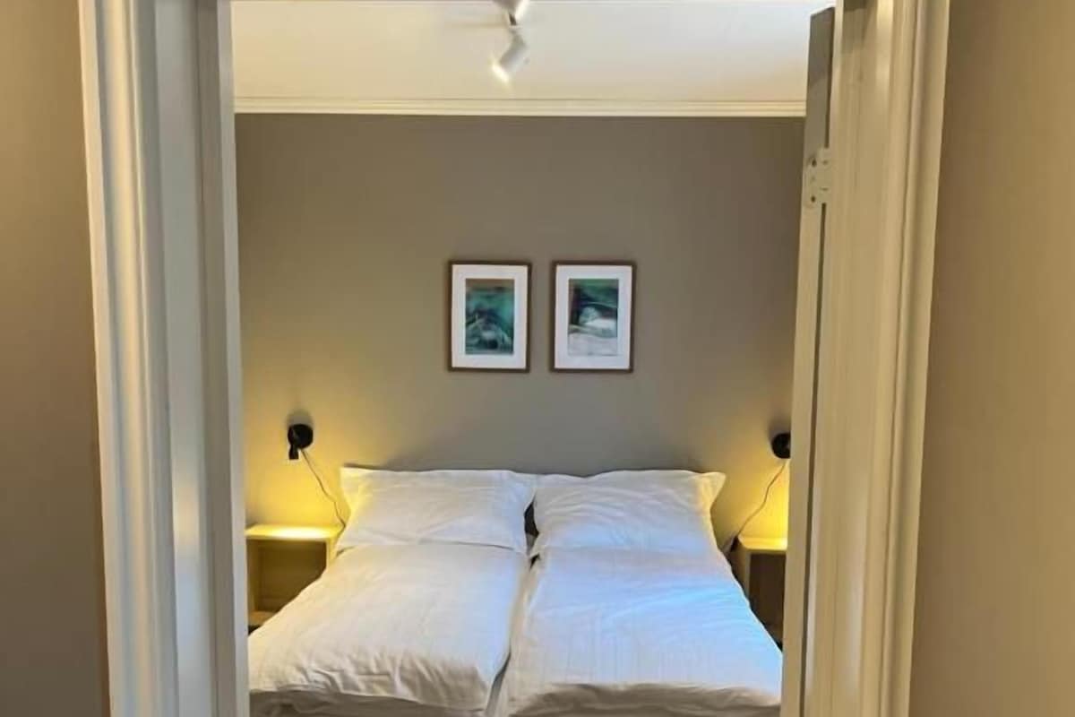 Uma porta estreita dá acesso a uma cama com roupa de cama branca, duas mesas de cabeceira fixadas na parede e duas fotografias emolduradas.
