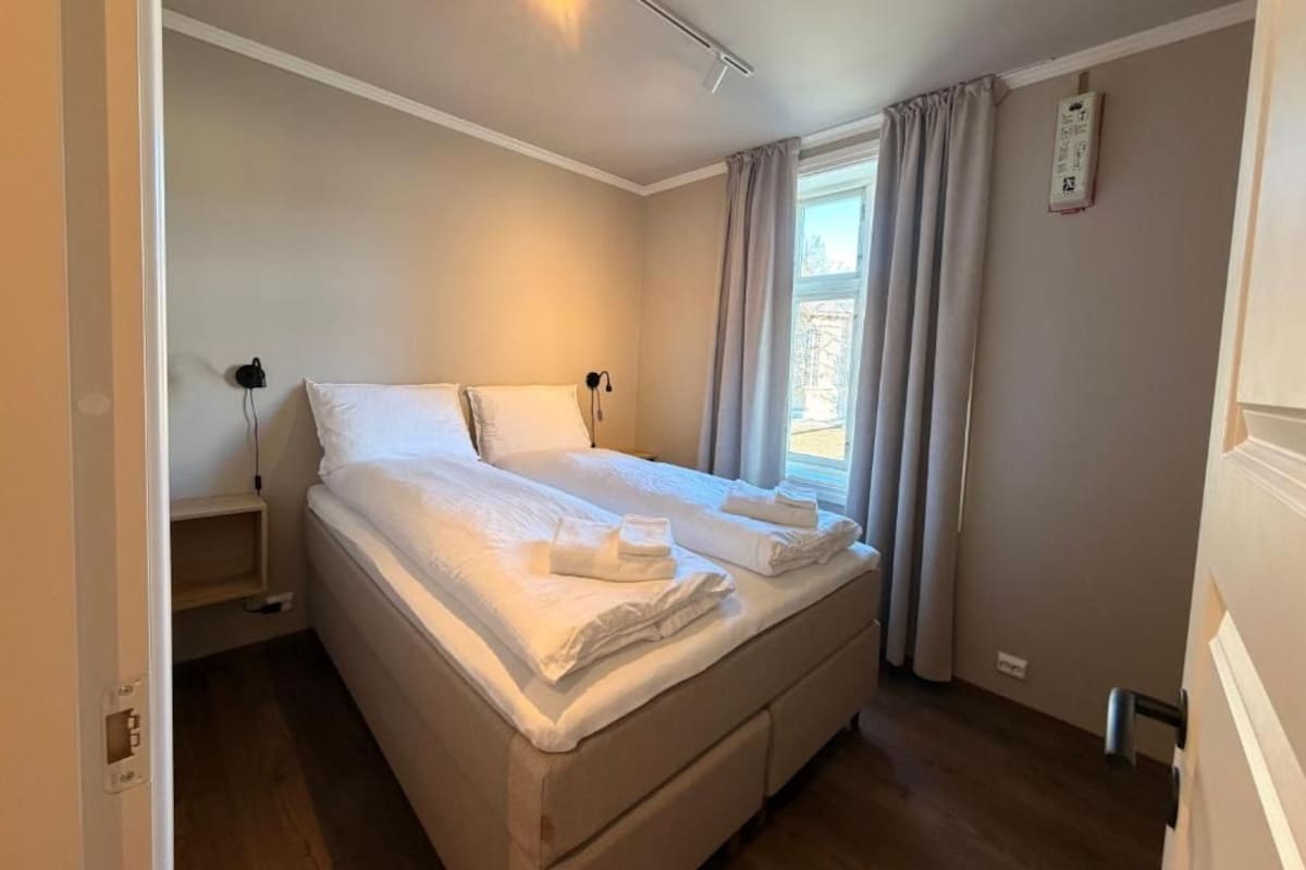 Cama bem arrumada com roupa de cama e toalhas brancas, mesa de cabeceira com luminária e janela com cortinas.