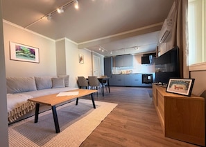 Uma sala de estar moderna com sofá, mesa de centro e cadeiras. Há uma cozinha ao fundo, com armários e uma televisão.