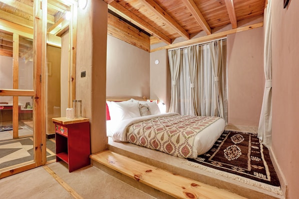 Quarto com teto de madeira, cama com roupa de cama estampada, mesa de cabeceira vermelha e janela com cortinas.