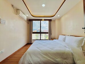 Quarto de hotel com uma cama grande, janela com cortinas, teto com iluminação embutida e aparelho de ar condicionado na parede.
