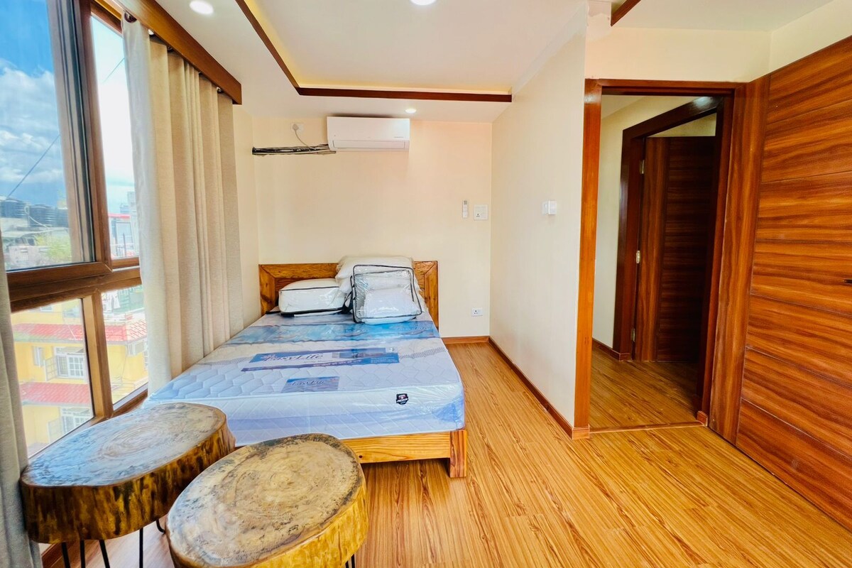 Quarto com cama, dois banquinhos, janela, aparelho de ar condicionado e porta de madeira.