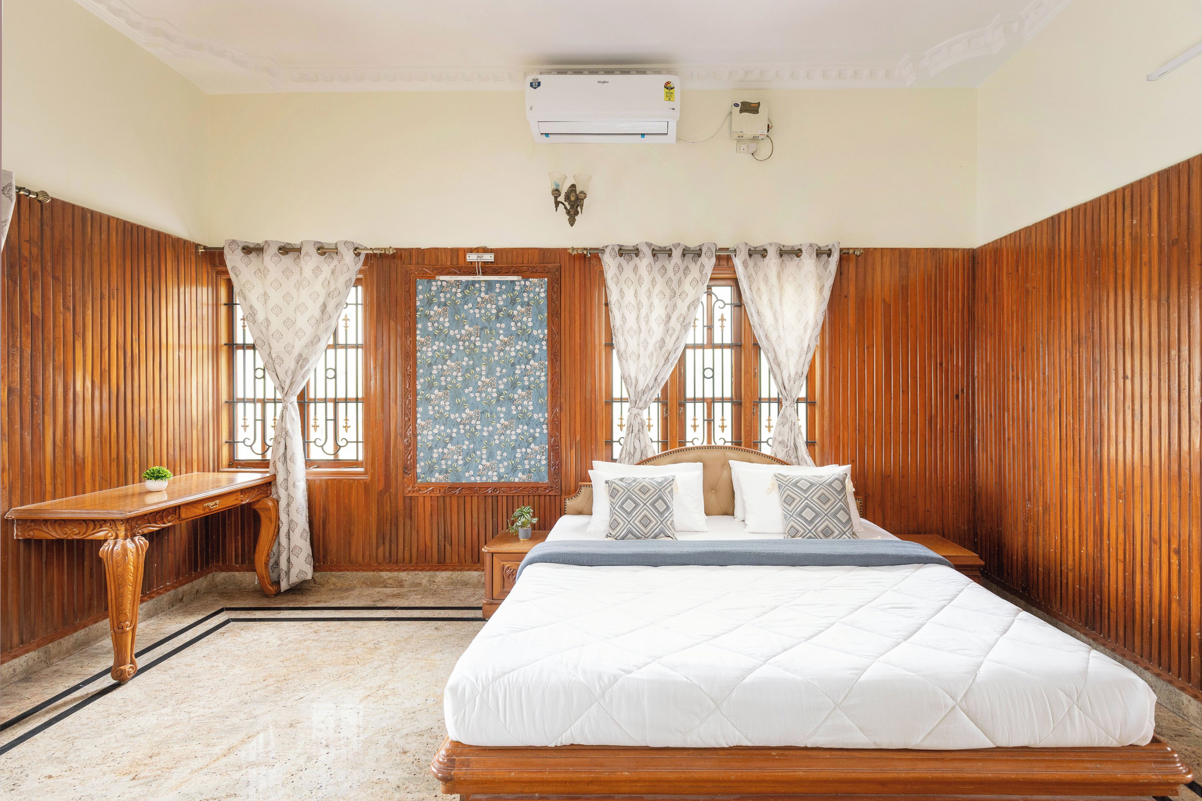 Quarto com parede de painel de madeira, cama com roupa de cama branca, uma mesinha com uma planta e uma janela com cortinas.