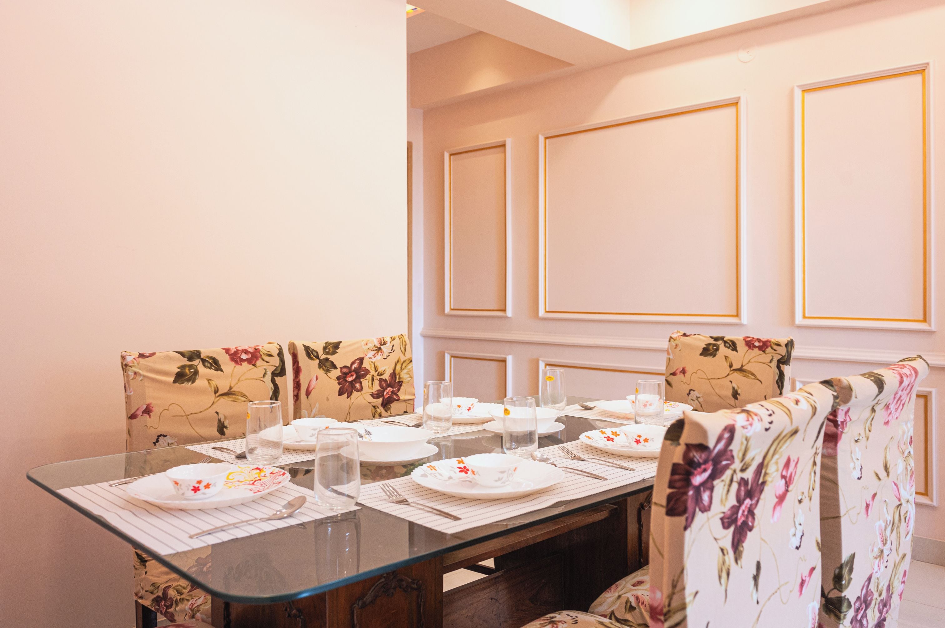 Uma área de jantar com mesa de vidro, cadeiras estofadas com motivos florais e uma parede branca com molduras decorativas.