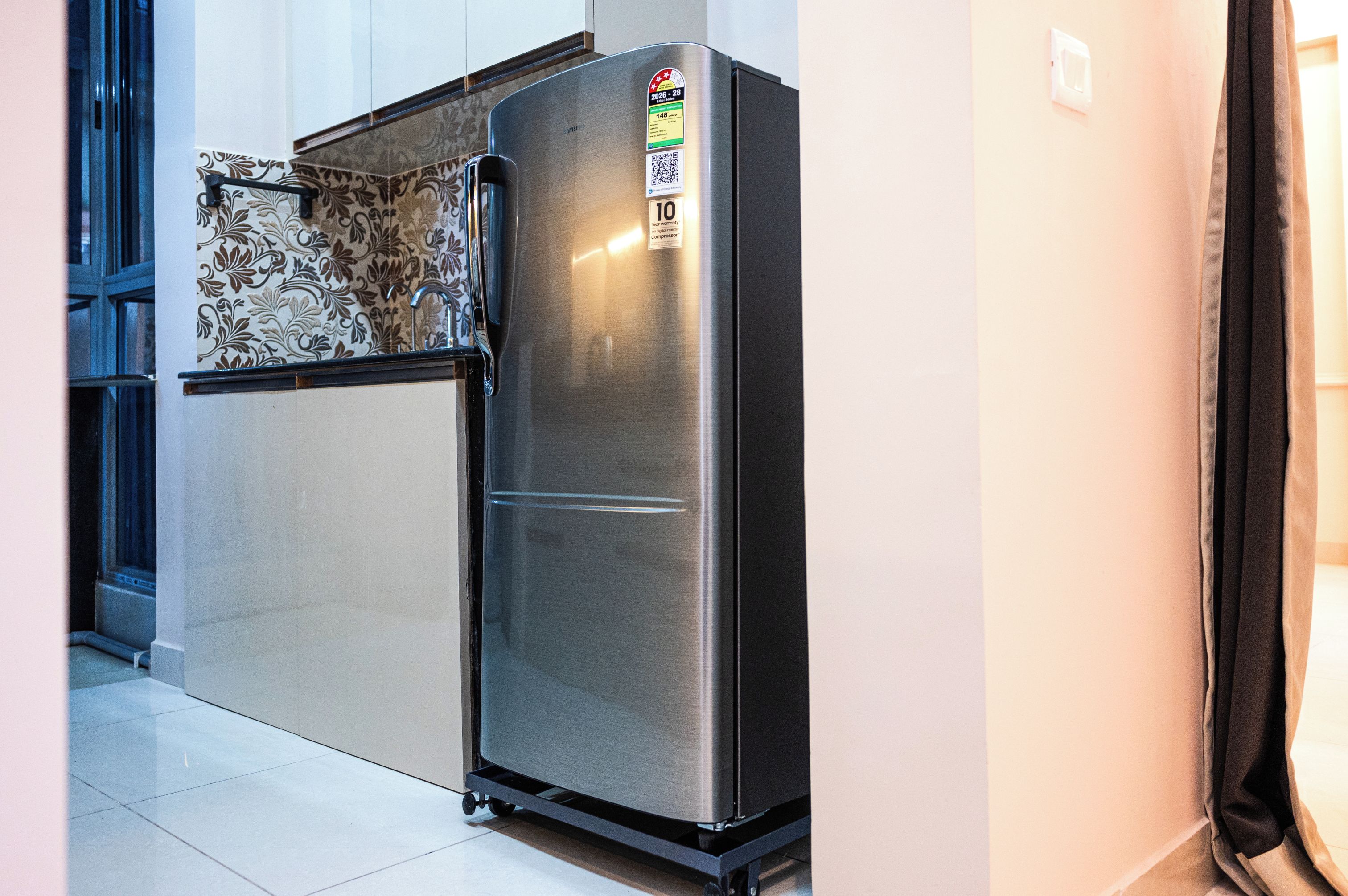 Uma geladeira de aço inoxidável com visor digital e adesivo de código de barras, colocada em um corredor com papel de parede estampado e uma porta com janela.