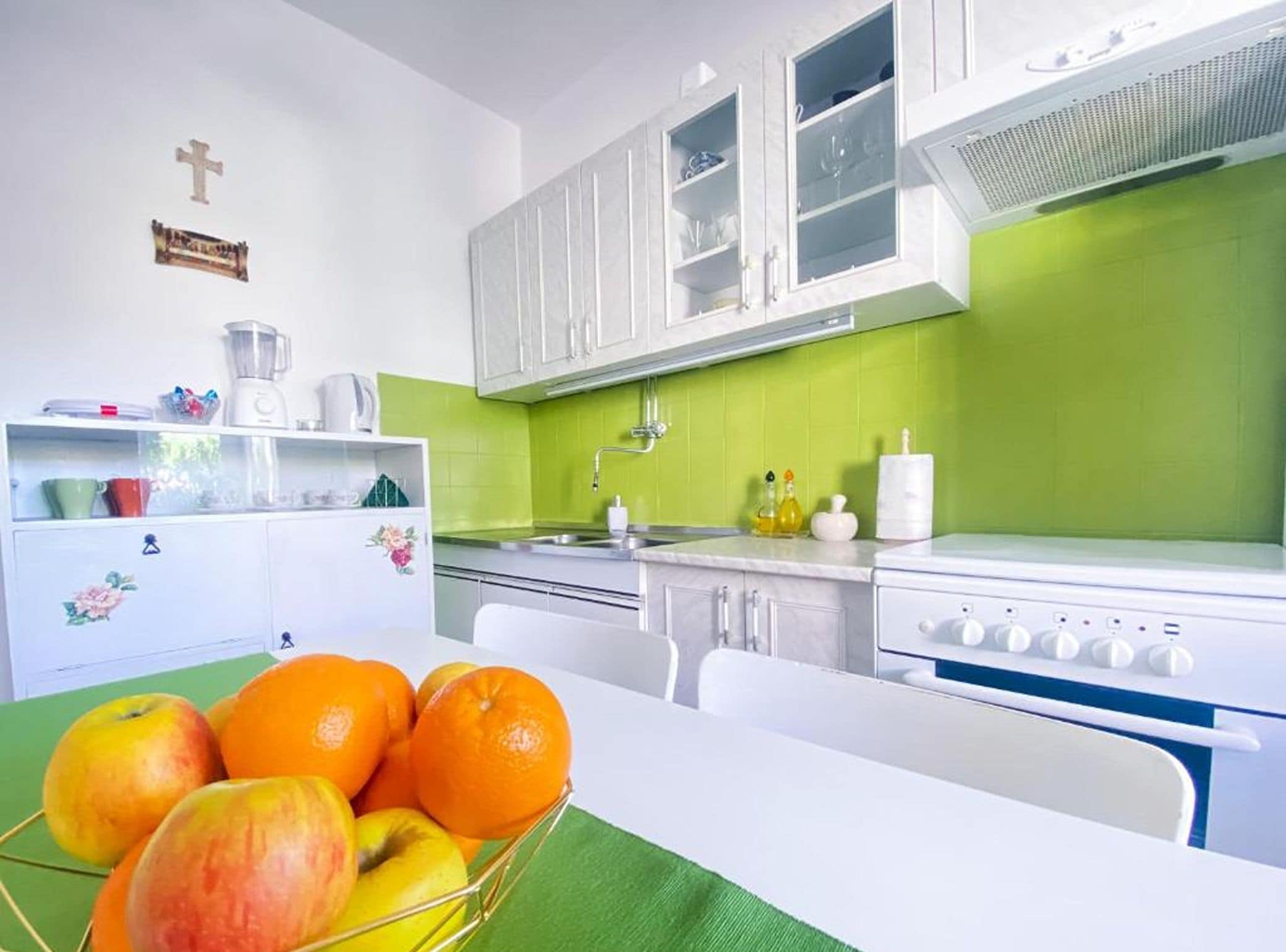 Cozinha com armários brancos, painel verde, pia, fogão e uma fruteira com maçãs e laranjas na bancada.