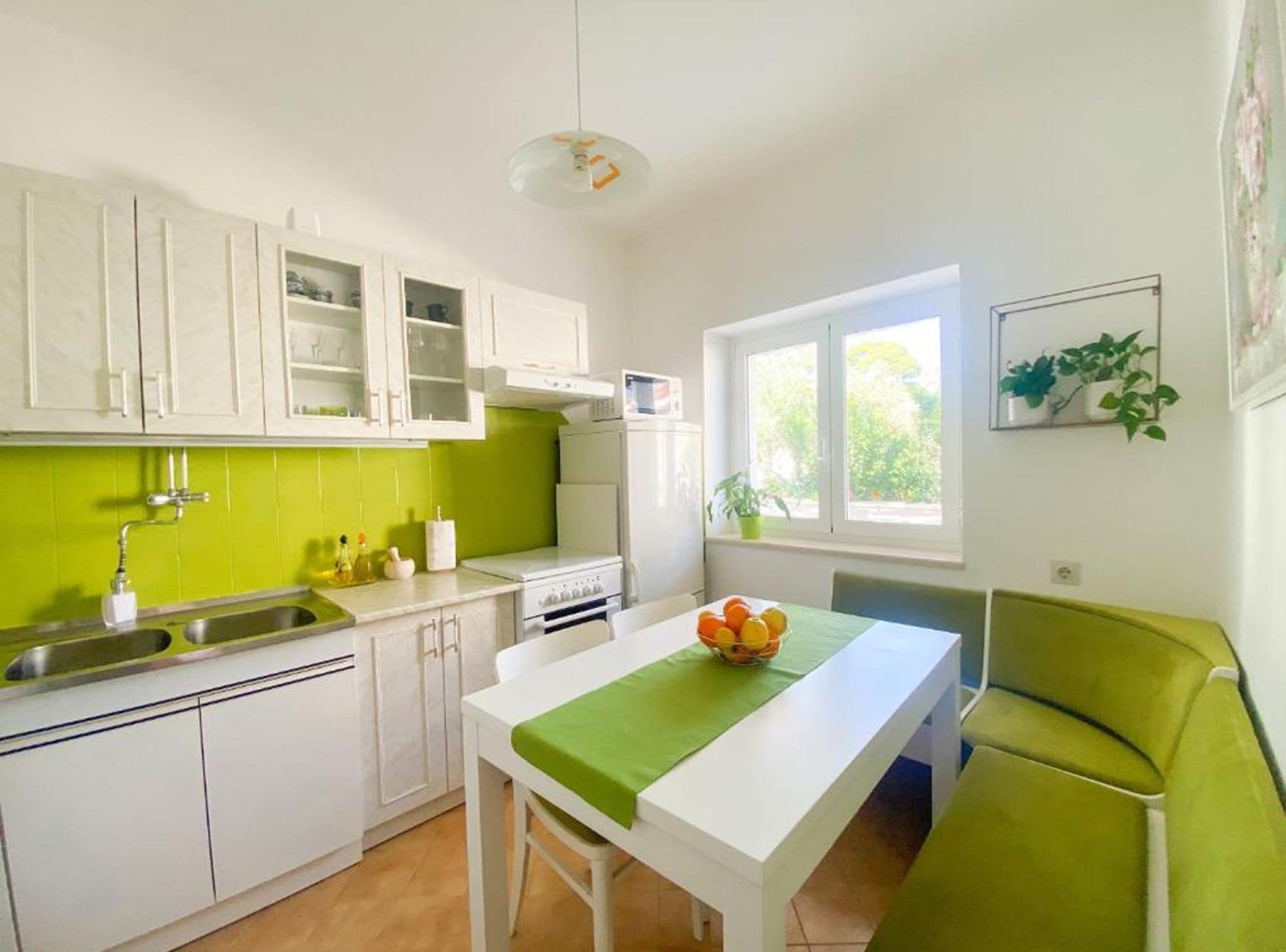 Cozinha compacta com armários brancos, painel verde, bancada branca, pia, fogão e janela com vista para vegetação.