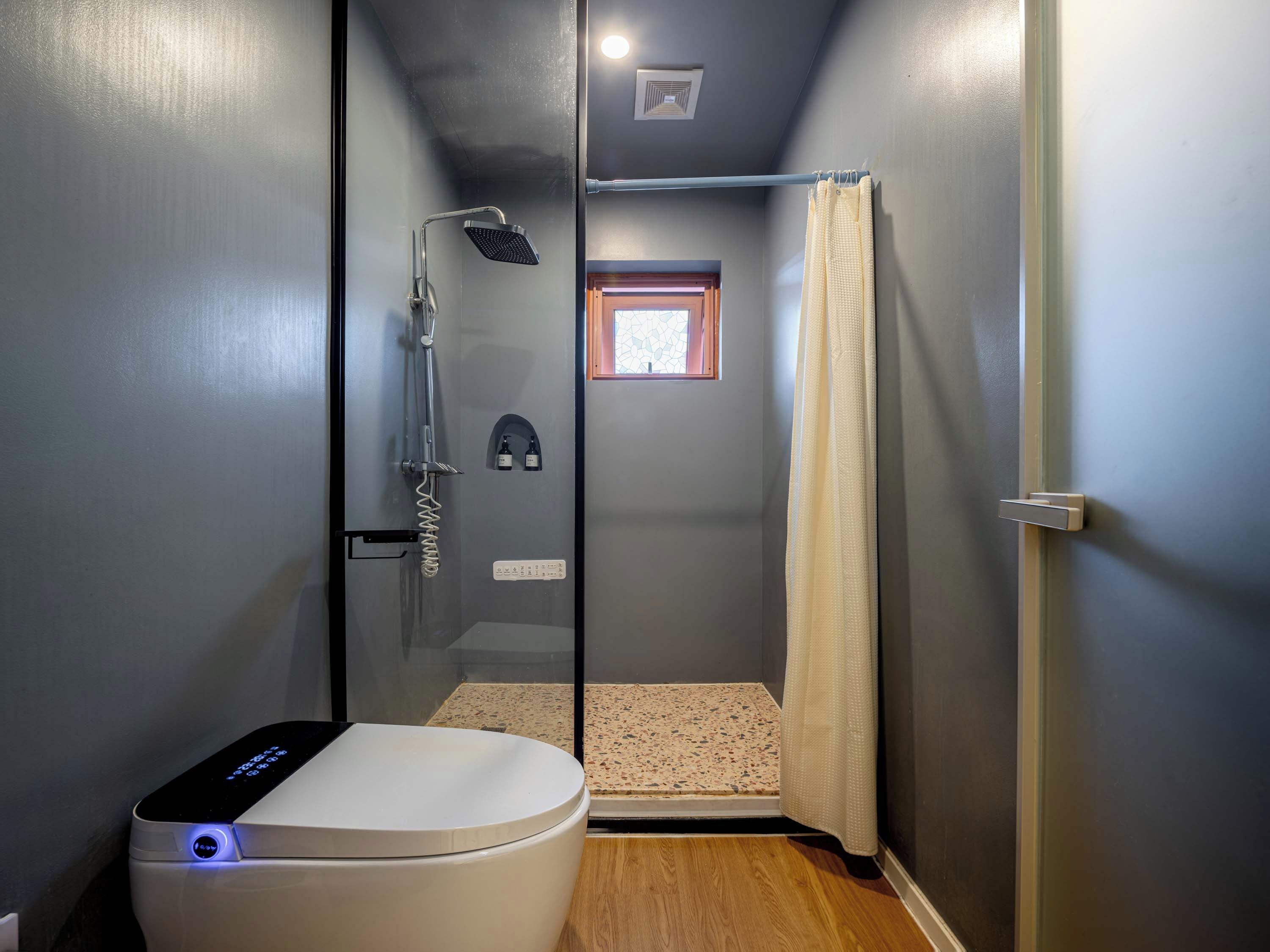 Casa de banho compacta com cabine de duche em vidro, sanita com painel digital e pequena janela.