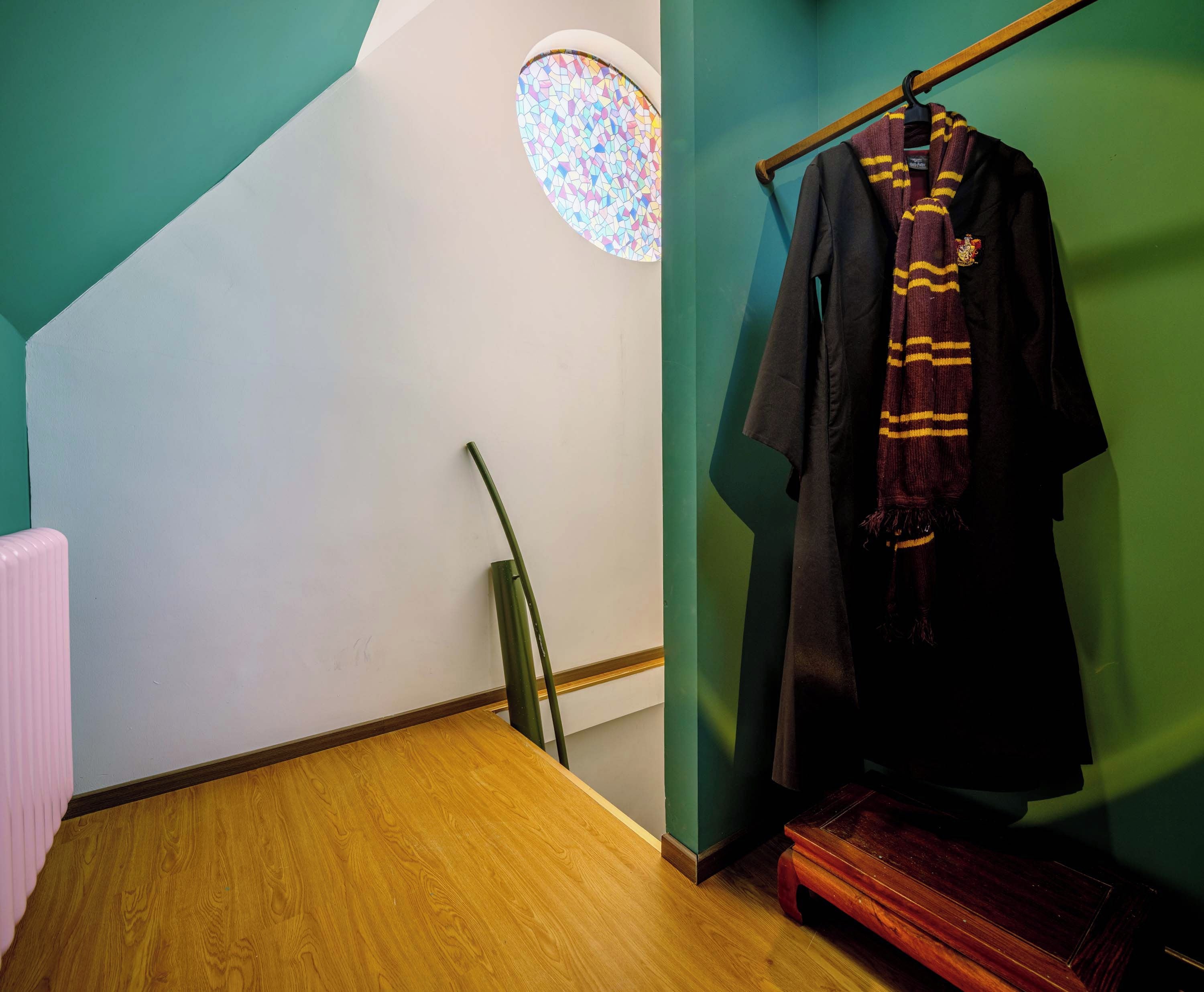 Um quarto com piso de madeira, parede verde, janela com design em vitral e casaco pendurado em um cabide.
