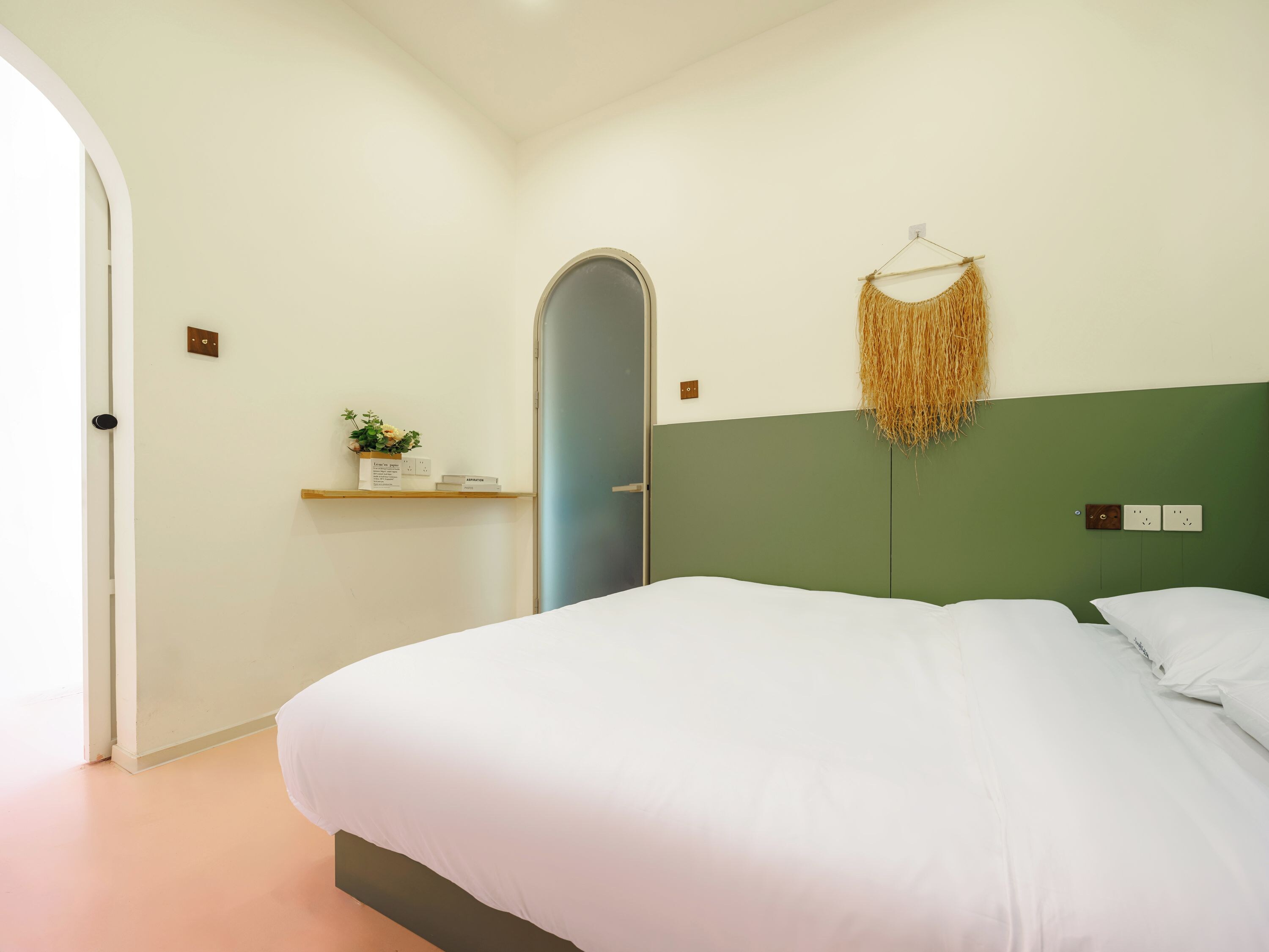 Quarto com cama, porta, prateleira com planta e luminária na parede.