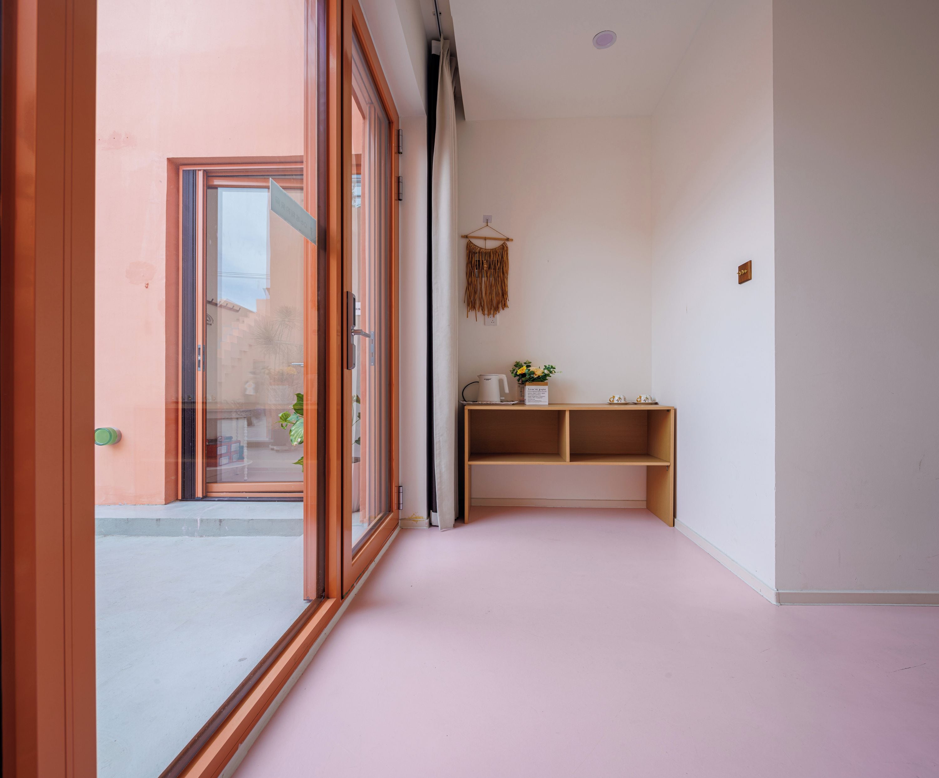Corredor com porta de vidro deslizante que dá para área externa, banco de madeira com mesa pequena e decoração de macramê pendente, além de parede branca com interruptor de luz.