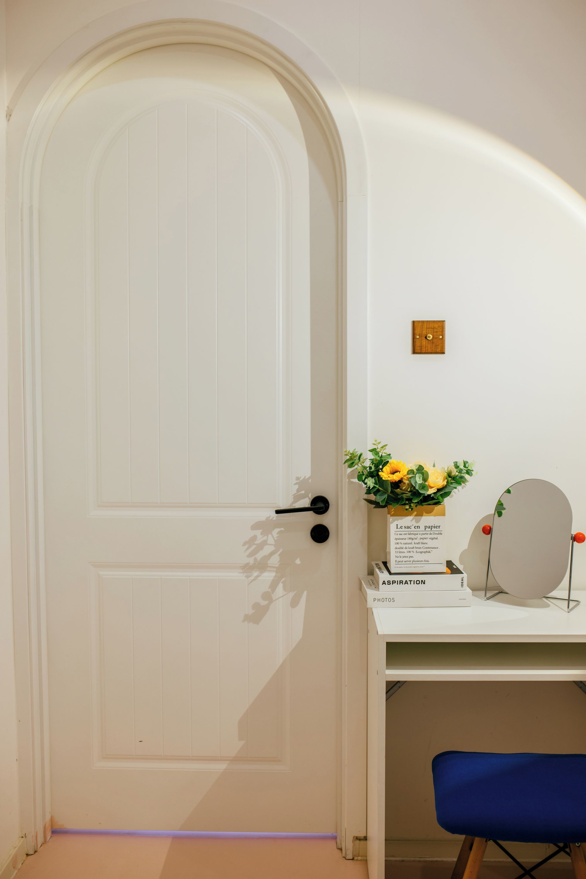 Uma porta branca com puxador de latão, uma pequena placa de madeira na parede, uma escrivaninha branca com espelho, um vaso com flores e uma cadeira azul.