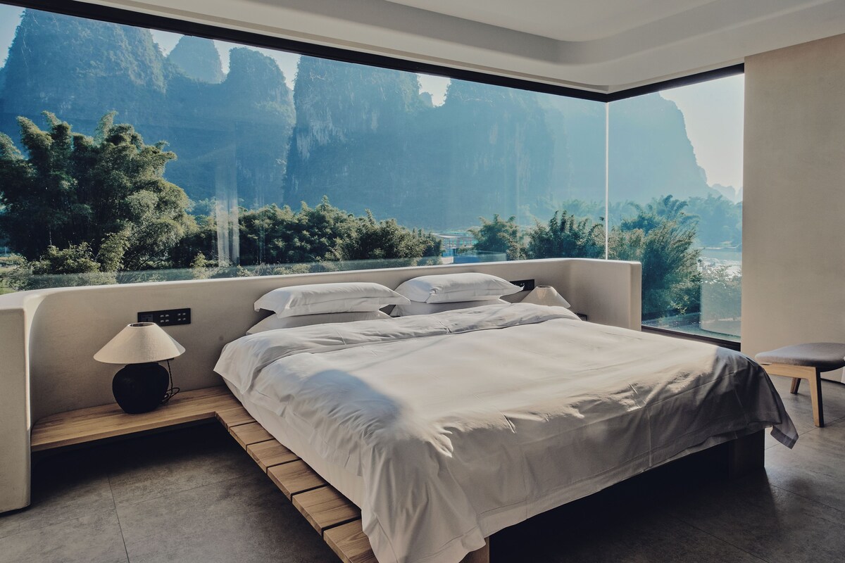 Quarto moderno com uma cama grande, mesa de cabeceira com abajur e vista para montanhas através de uma janela ampla.
