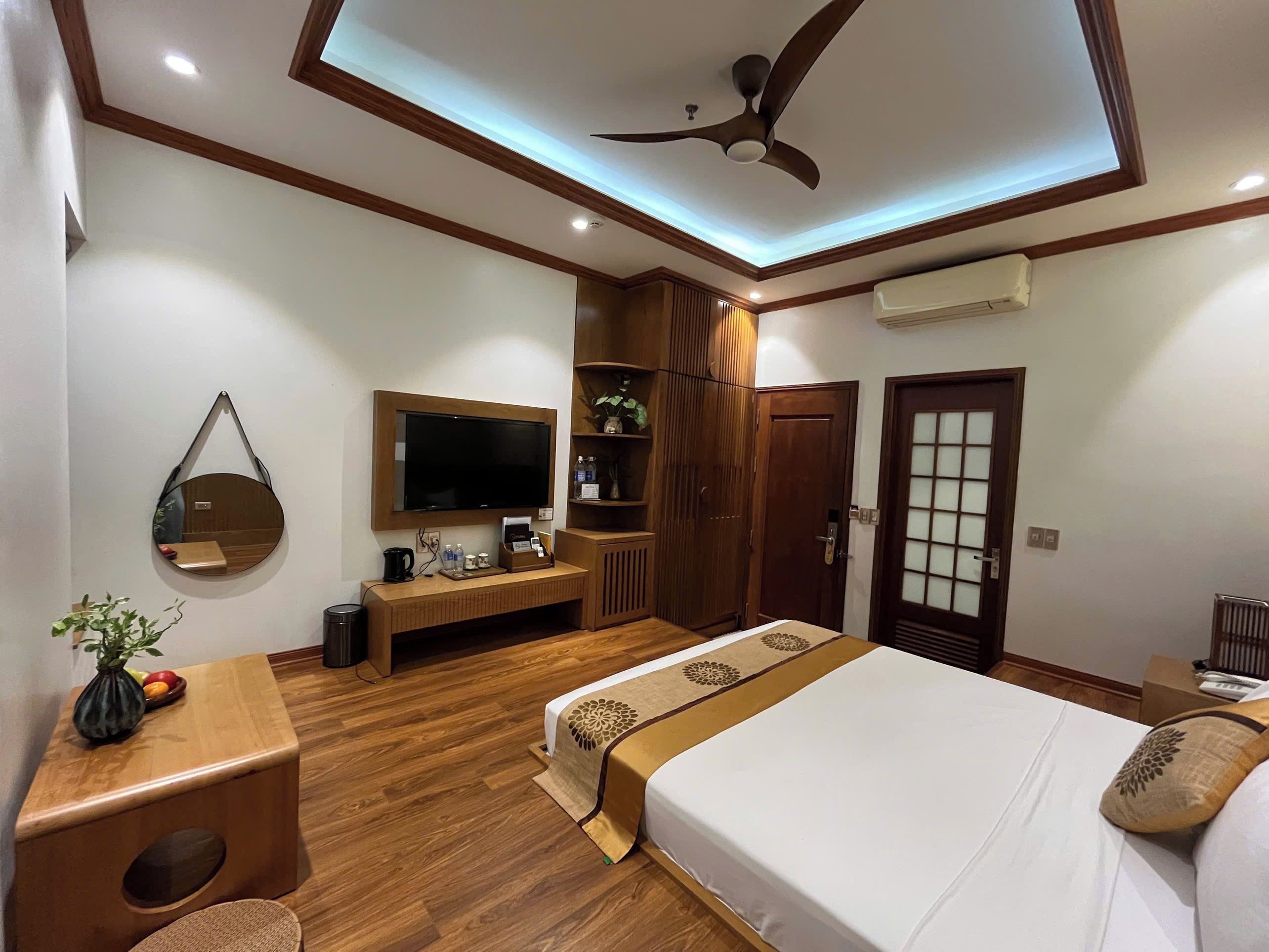 Quarto de hotel com cama, televisão, escrivaninha com espelho, ventoinha no teto e guarda-roupa.