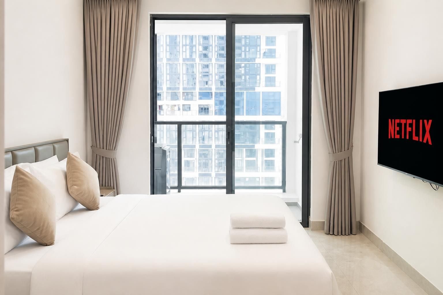 Quarto de hotel com cama, cortinas, uma televisão exibindo o logotipo da Netflix e vista para edifícios através de uma janela grande.