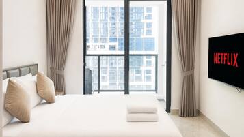 Quarto de hotel com cama, cortinas, uma televisão exibindo o logotipo da Netflix e vista para edifícios através de uma janela grande.
