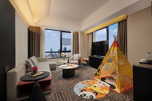 Quarto de hotel moderno com sofá, poltronas, mesa de centro, TV e uma tenda infantil.