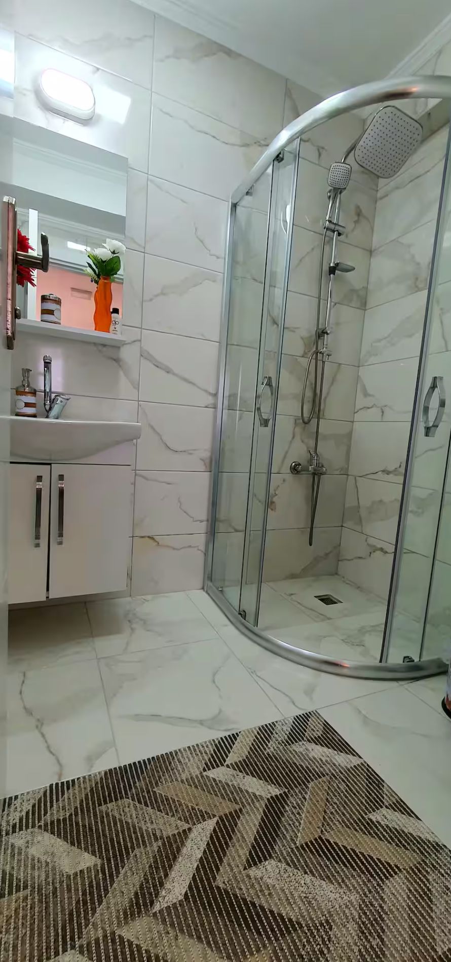 Casa de banho com cabine de duche em vidro, parede revestida a mármore branco e lavatório branco com espelho em cima.