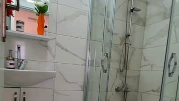 Casa de banho com cabine de duche em vidro, parede revestida a mármore branco e lavatório branco com espelho em cima.