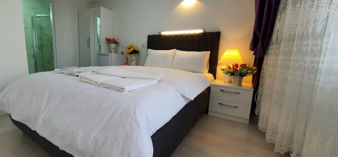 Cama bem arrumada com roupa de cama branca, abajur e uma mesinha de cabeceira com um vaso de flores.
